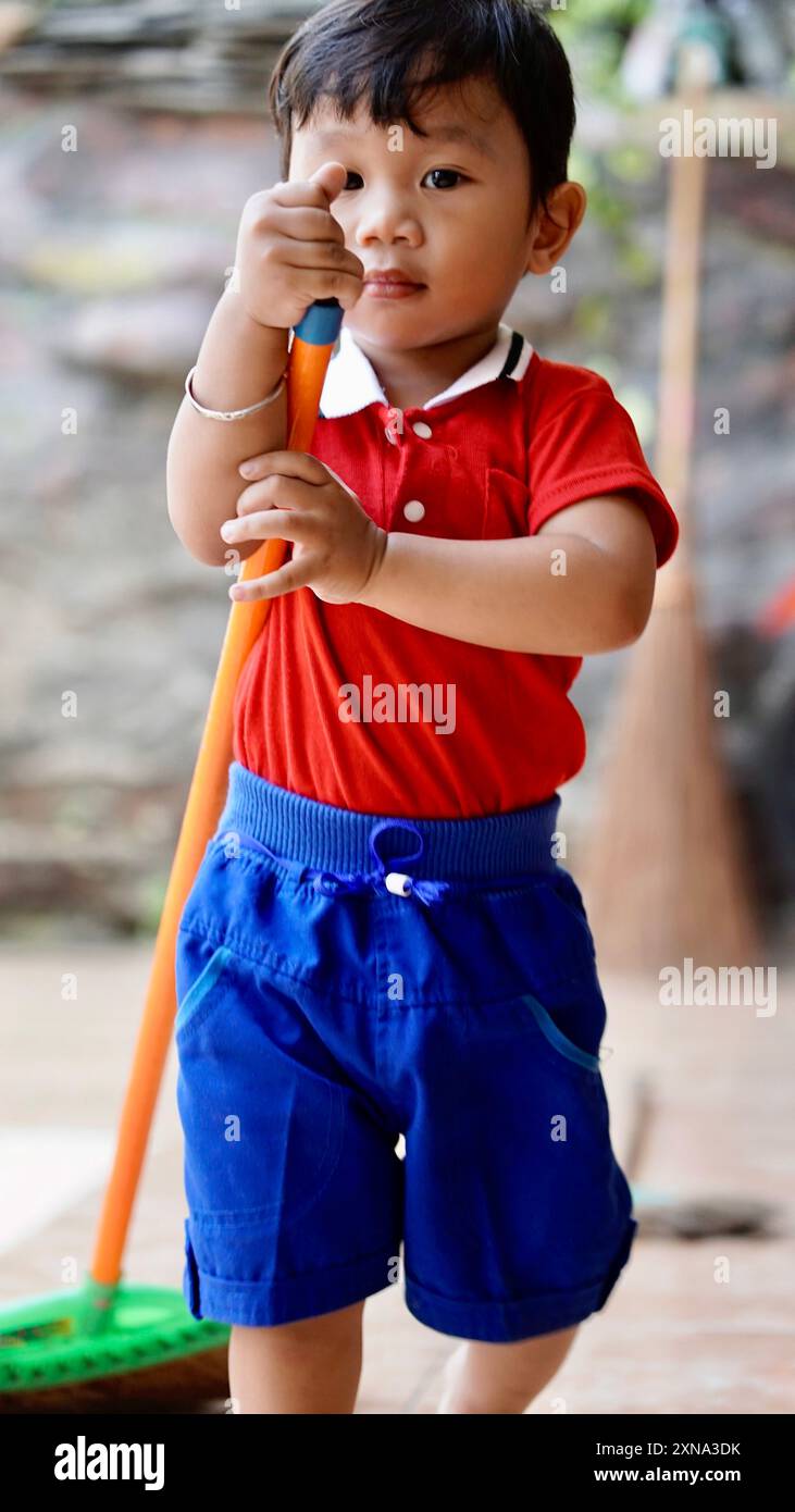 A young boy in a red shirt and blue shorts is holding a green broom ...