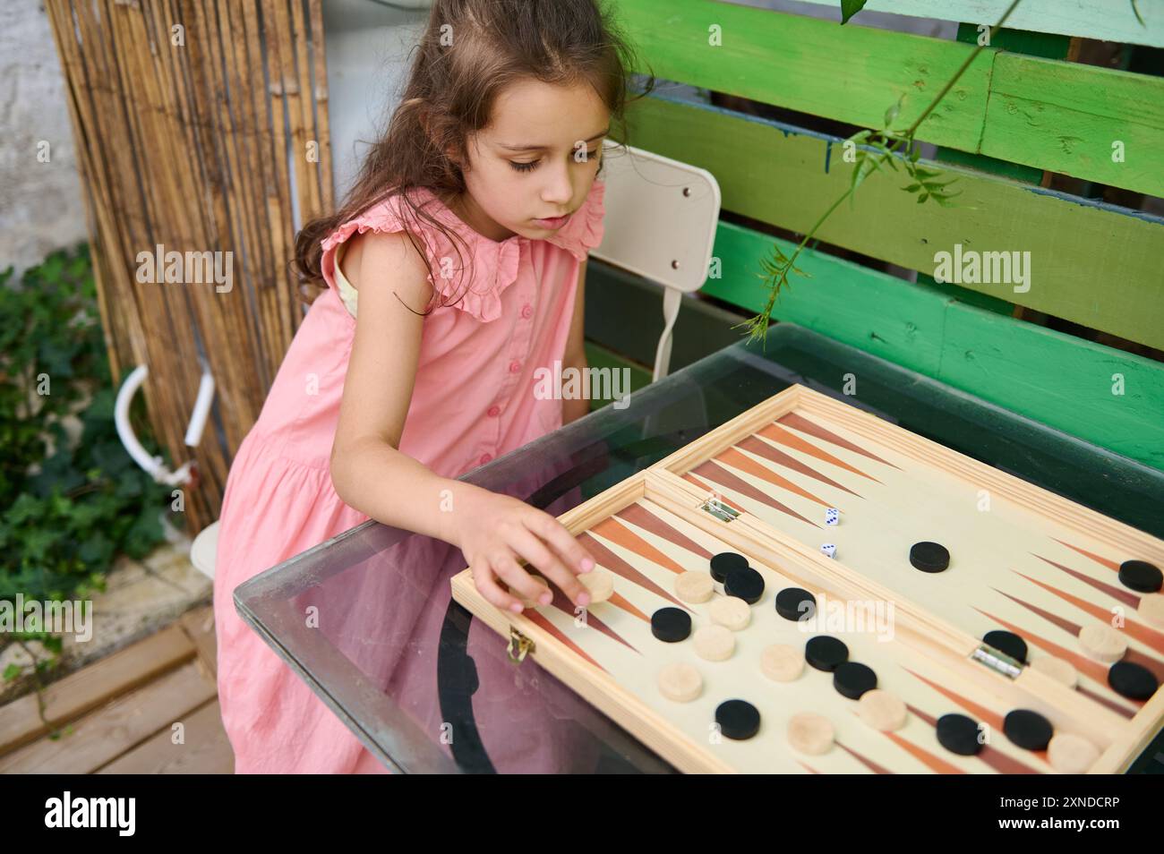 Young girl in a pink dress playing backgammon at home. Focused child enjoys indoor activity with ...