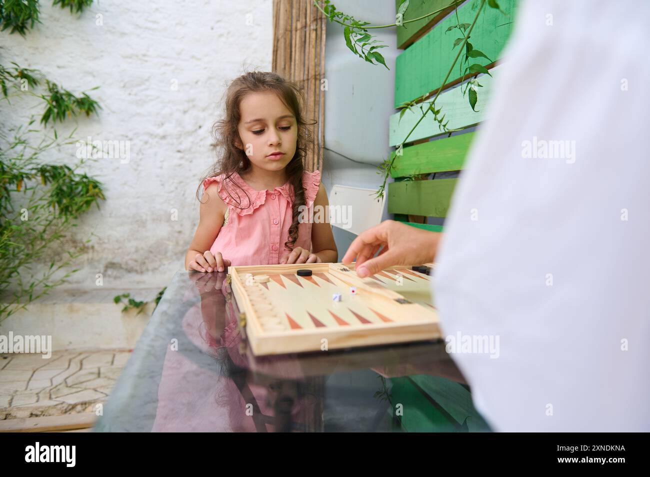 Young girl in a pink dress concentrating on learning to play backgammon ...