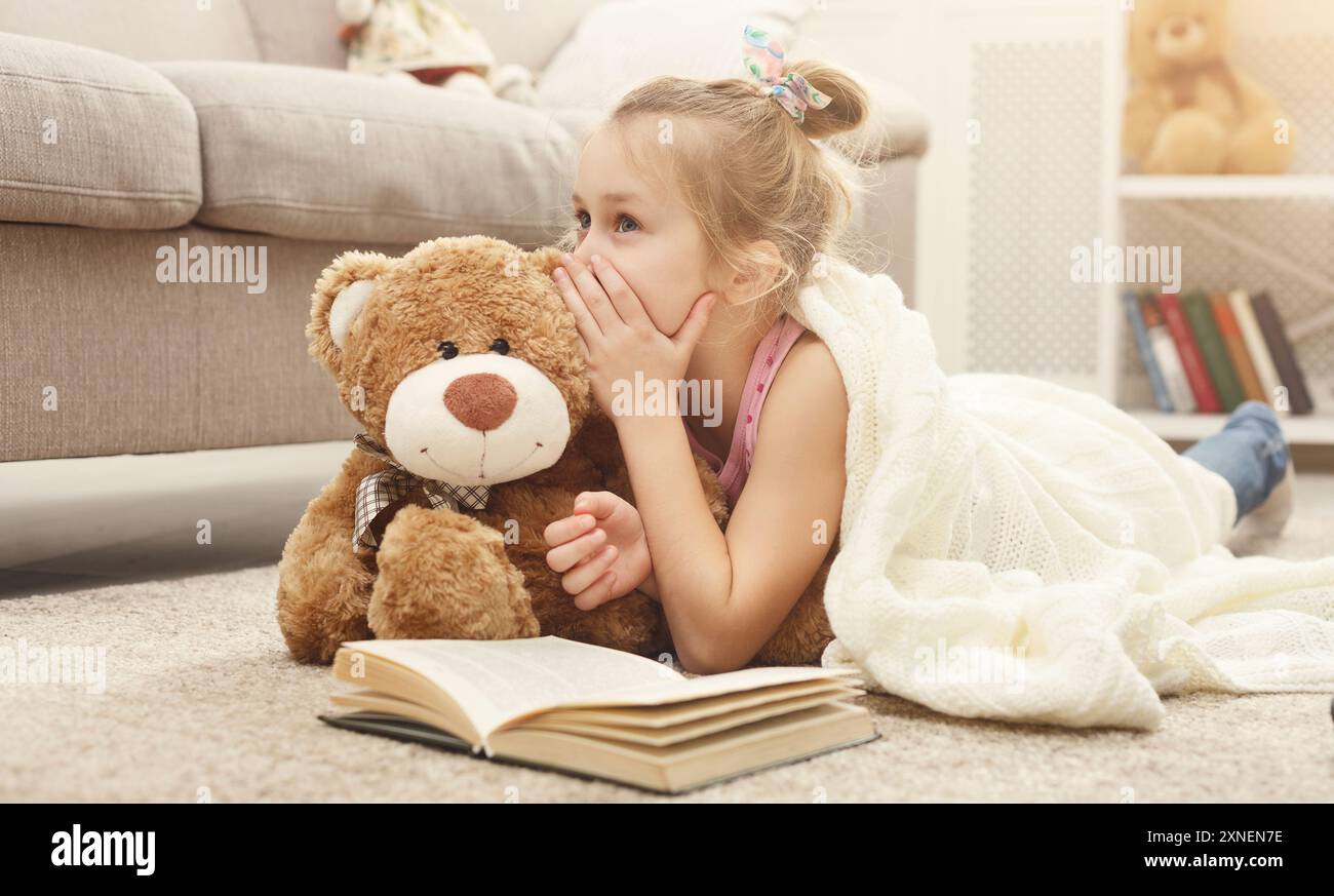 Happy little female child and her teddy bear reading book on the floor ...