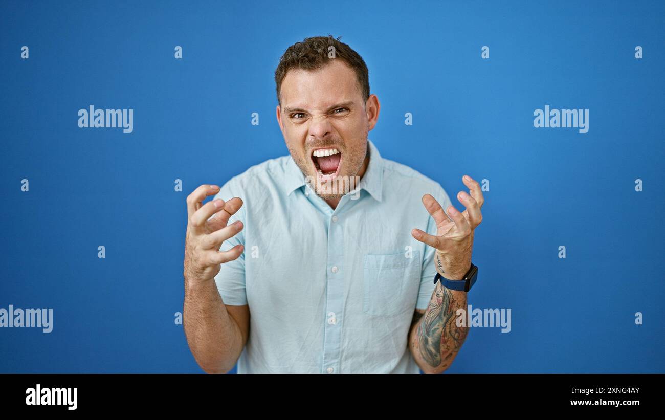 A frustrated young man with a beard expressing anger in front of a blue ...