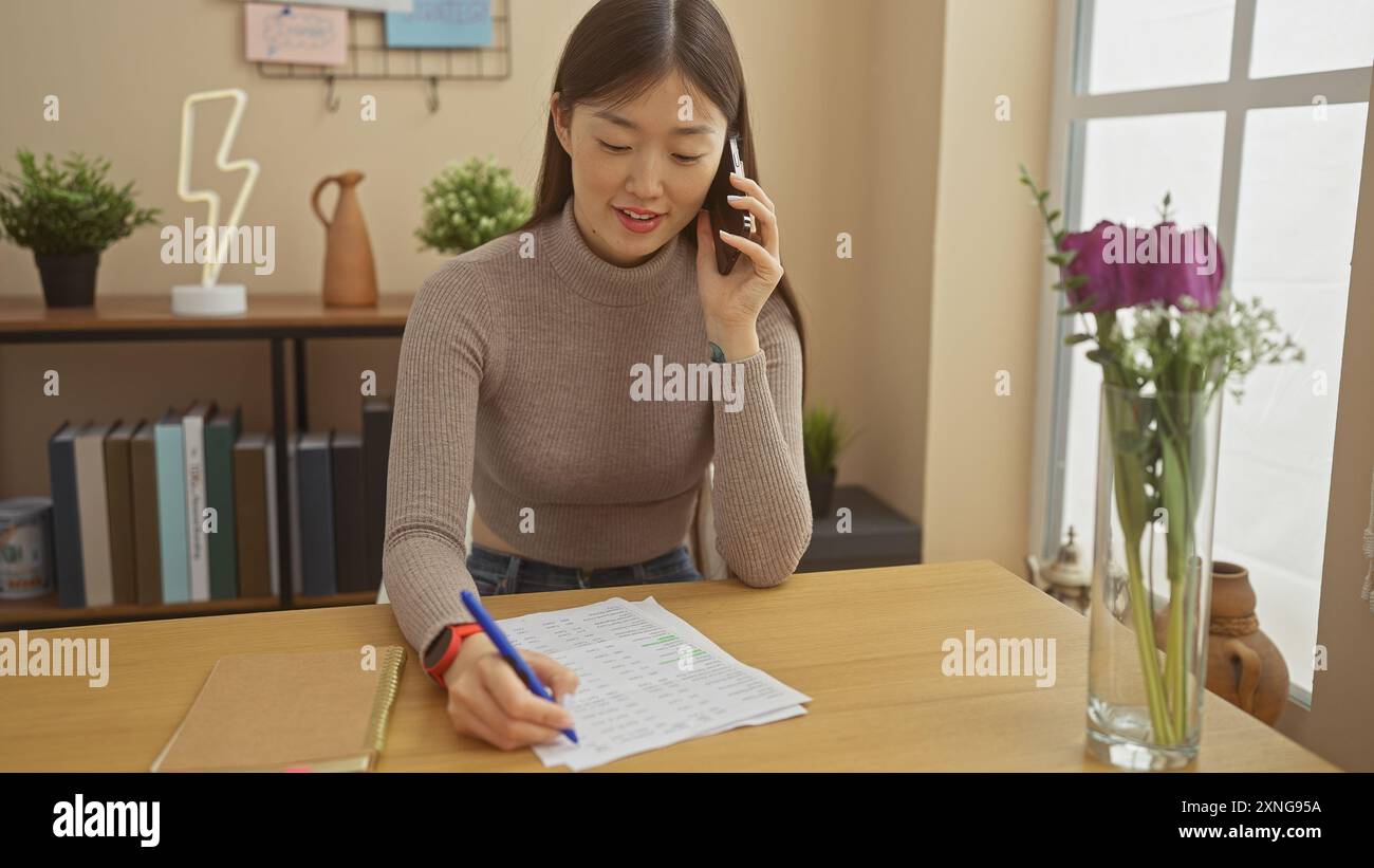 A young asian woman talks on the phone while writing notes at a wooden ...
