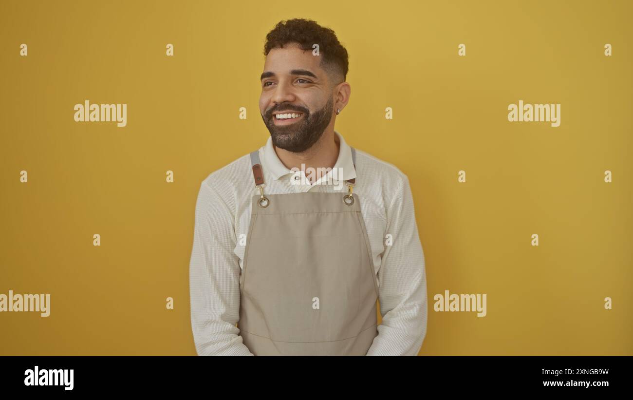 A cheerful young hispanic man with a beard wearing an apron stands ...