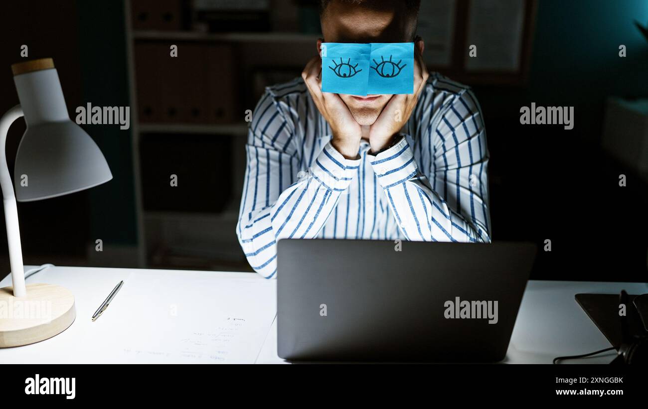 A young man holds a sticky note with drawn eyes over his face, in a ...