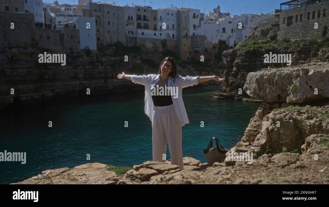A young hispanic woman joyfully poses on the rocky cliffs of polignano ...
