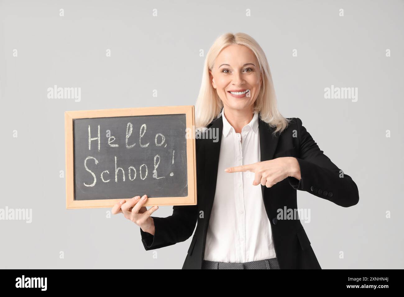 Mature female teacher pointing at chalkboard with text HELLO SCHOOL on ...