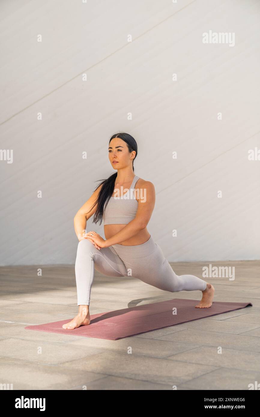 A woman practicing yoga in a lunge pose on a mat indoors with a light ...