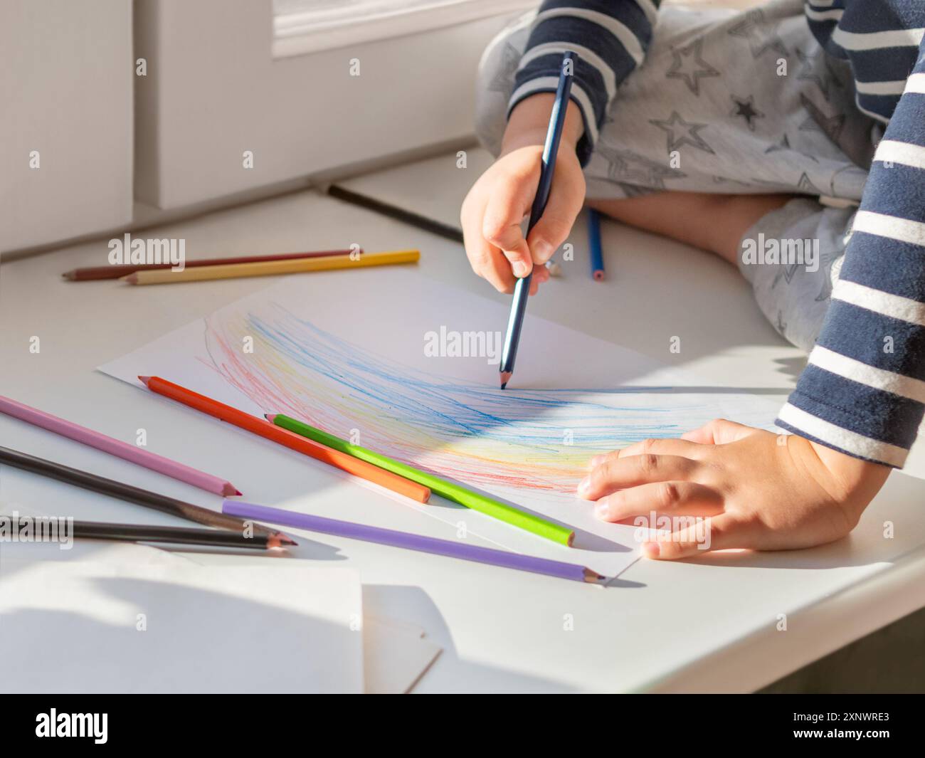 Little boy sits on window sill and draws rainbow with colored pencils ...
