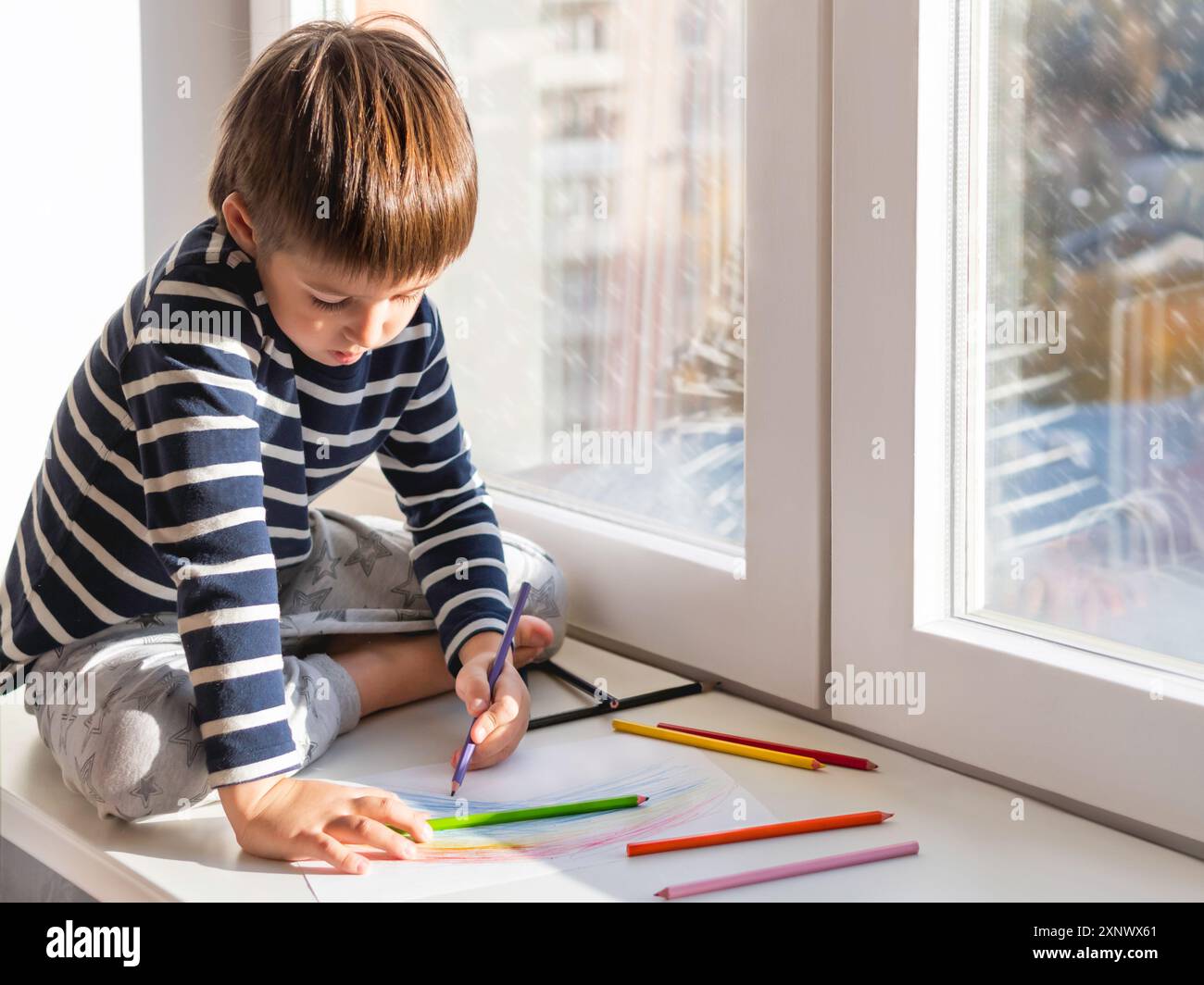 Left-handed boy sits on window sill and draws rainbow with colored ...