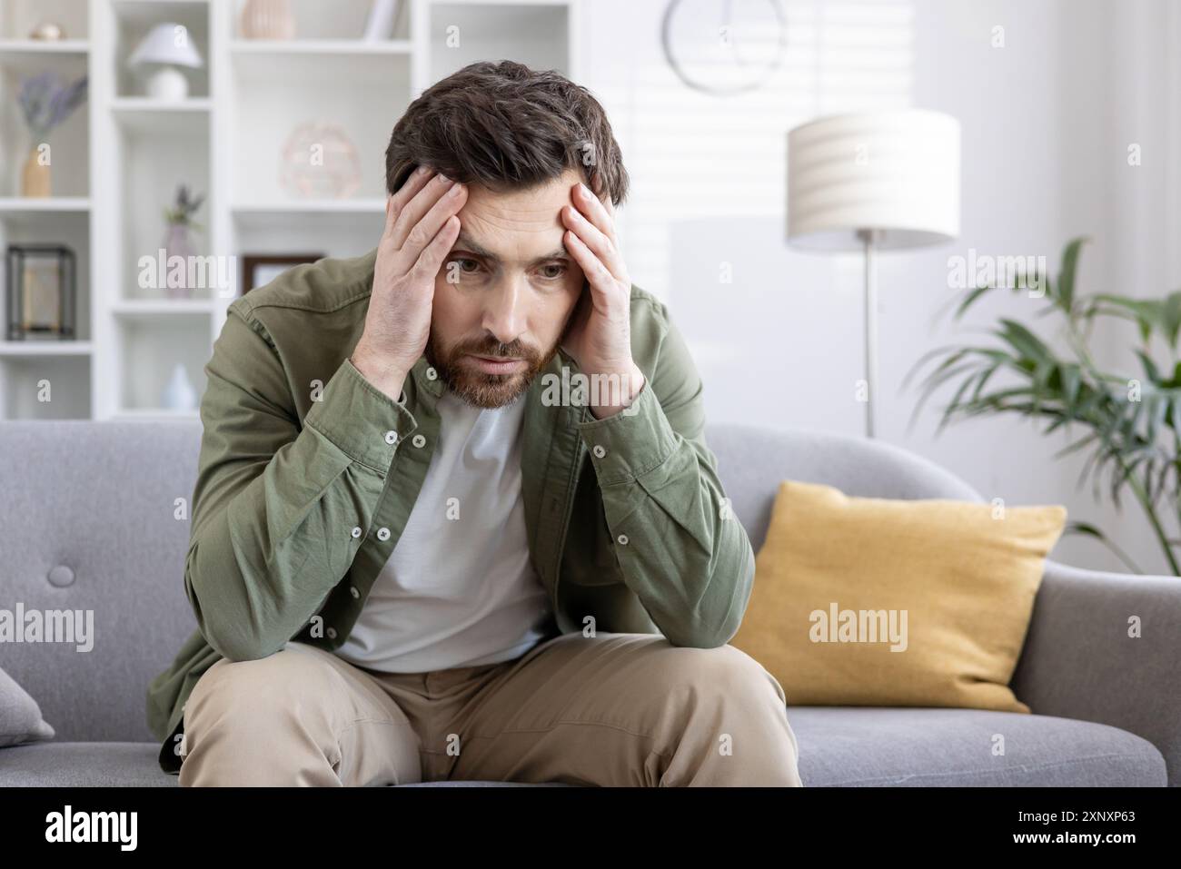 Stressed man sitting on sofa holding his head, feeling overwhelmed and ...
