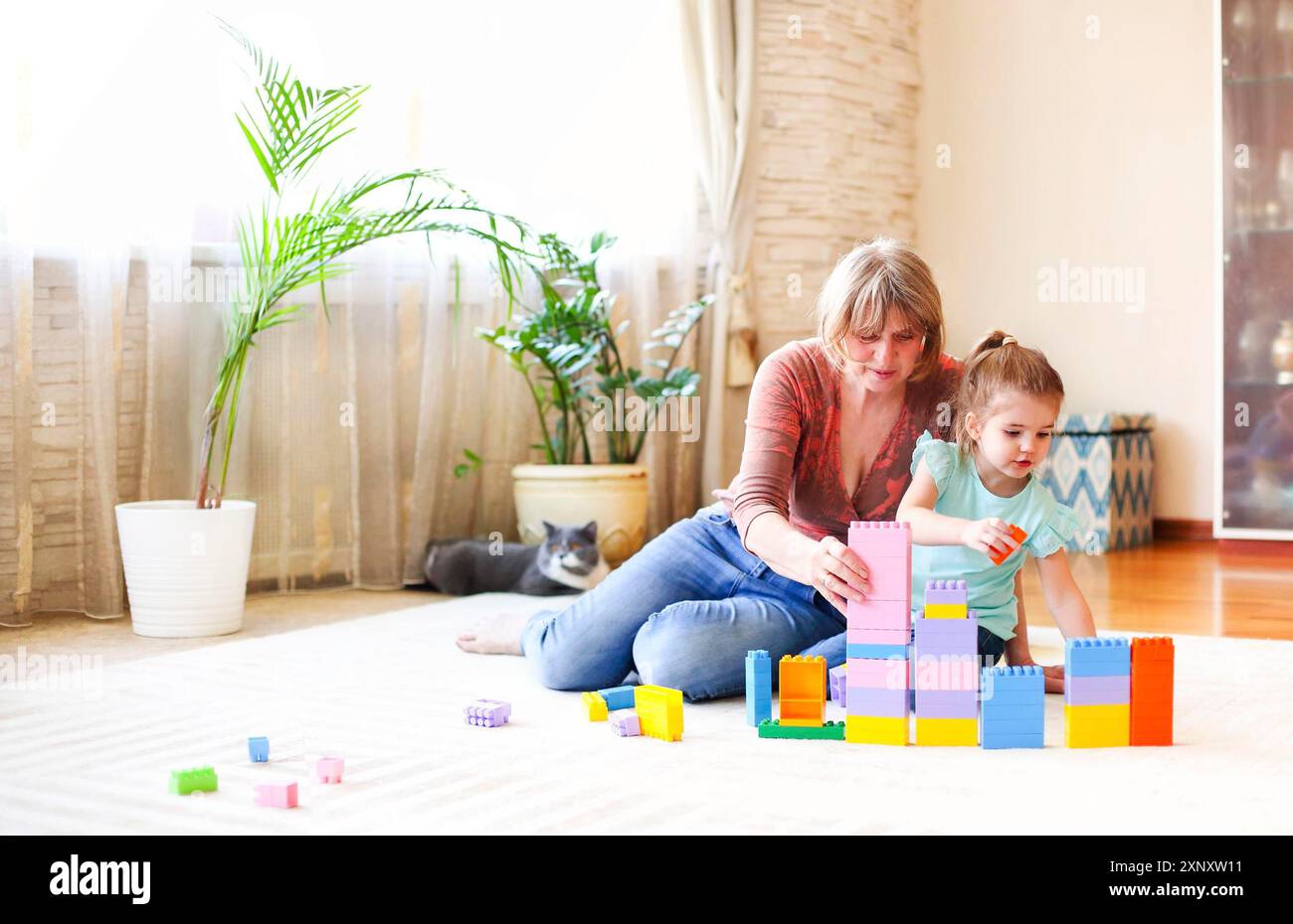 Cheerful mature woman watching little girl building tower from bricks ...