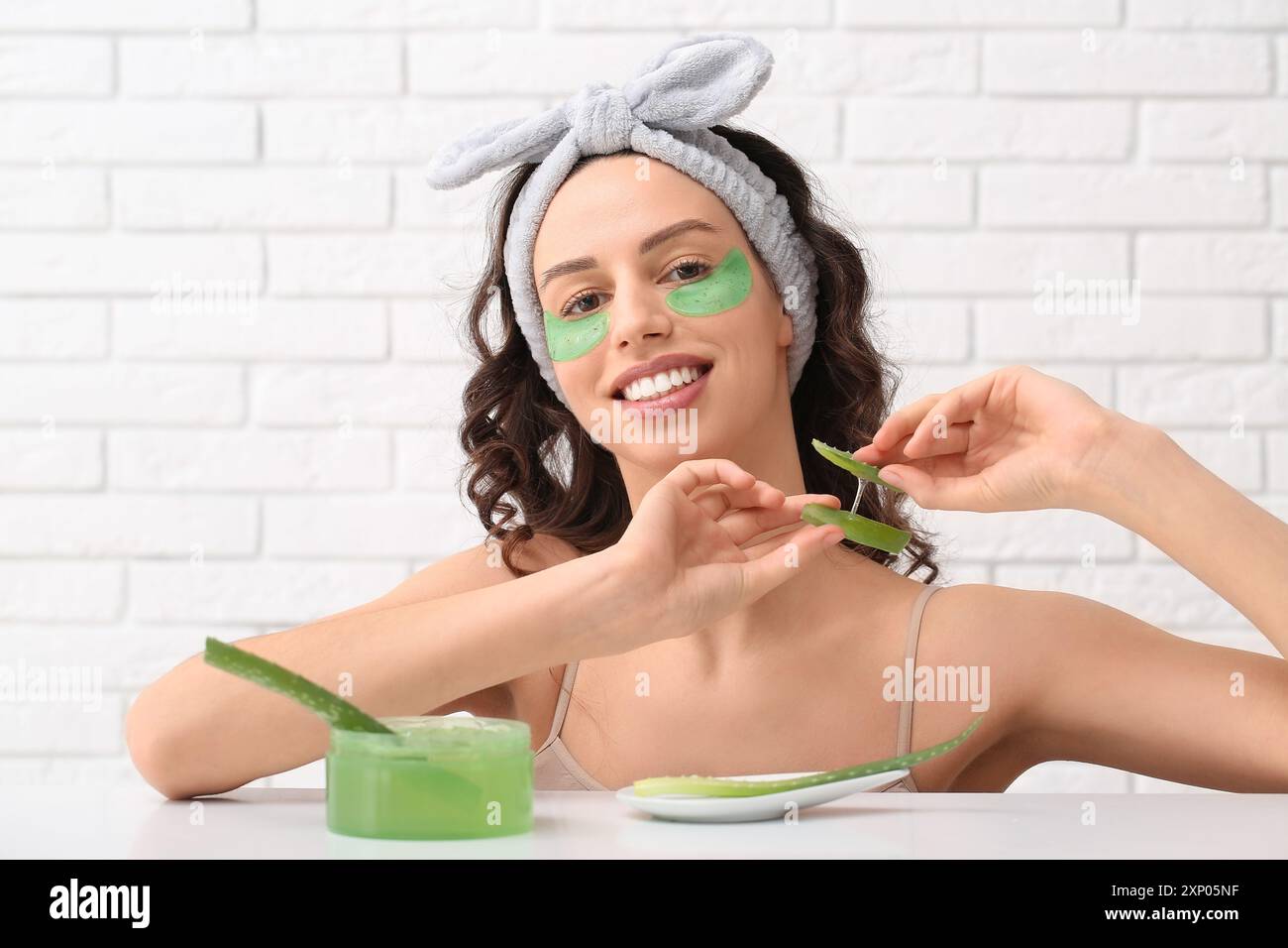 Beautiful young woman applying aloe vera leaf on white brick background ...