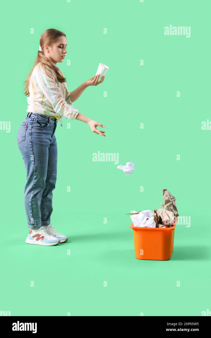 Young woman throwing paper into trash bin on green background. Waste ...