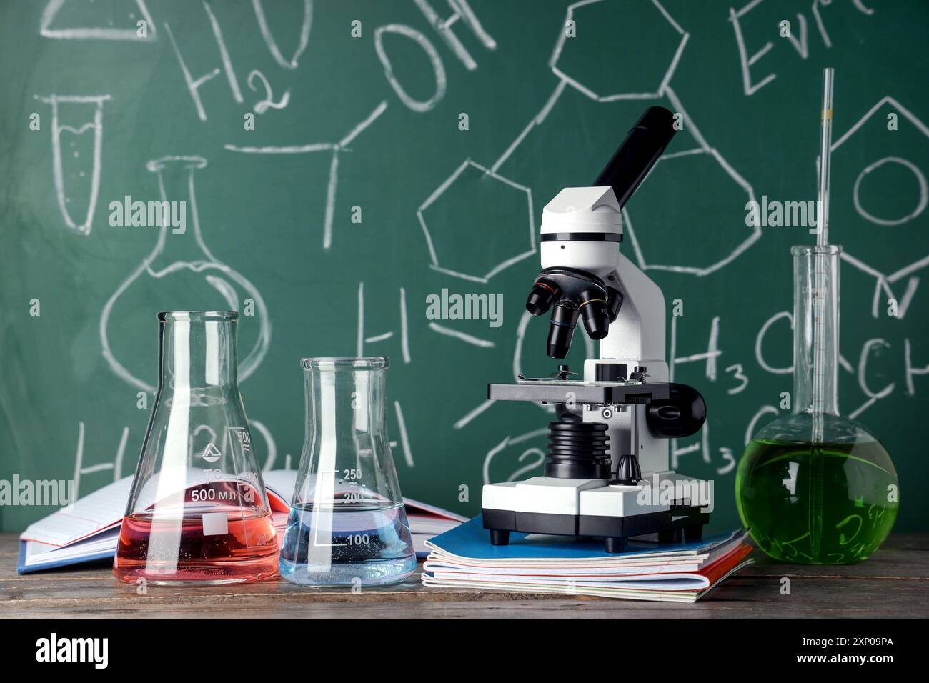 Modern microscope and flasks on wooden table near chalkboard with ...