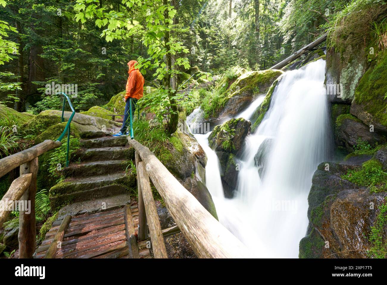 A person in an orange jacket stands on wooden steps beside a cascading ...