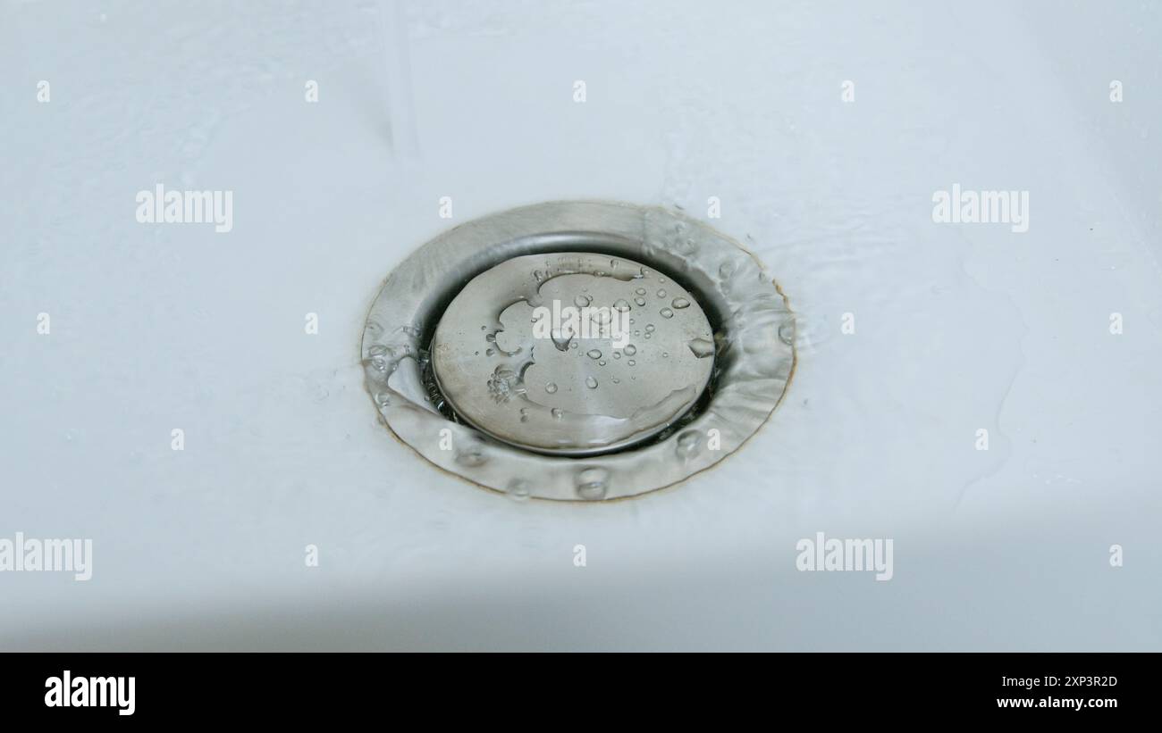 A close-up view of a stainless steel drain in a white sink. Water is ...