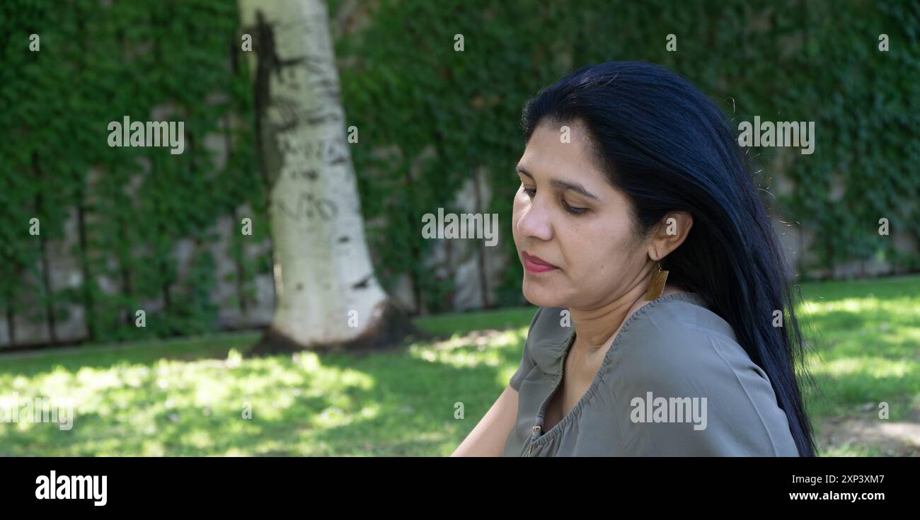 A woman with long hair is sitting in a park. She is wearing a green ...