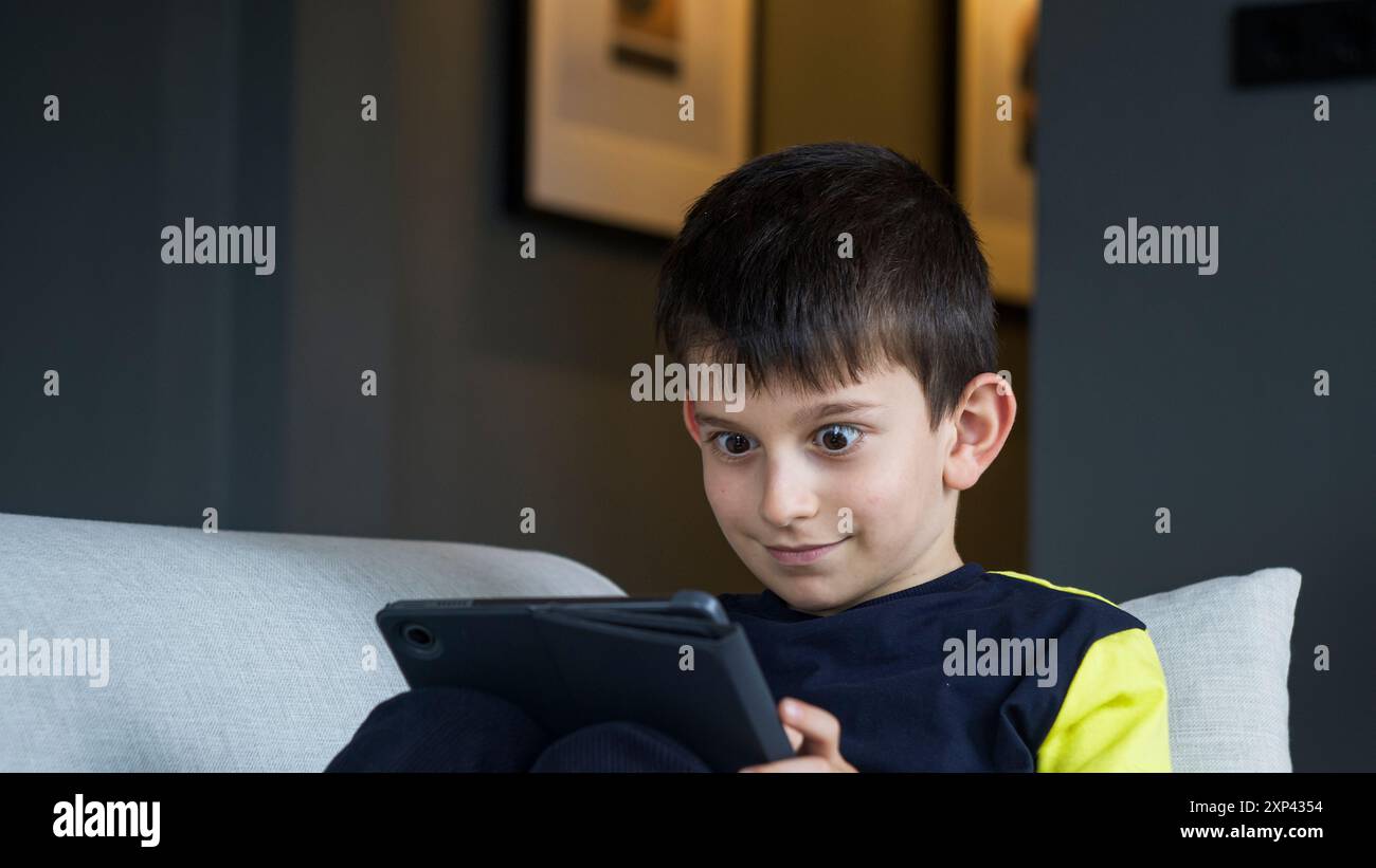 A young boy is sitting on a couch, intensely focused on a tablet he ...