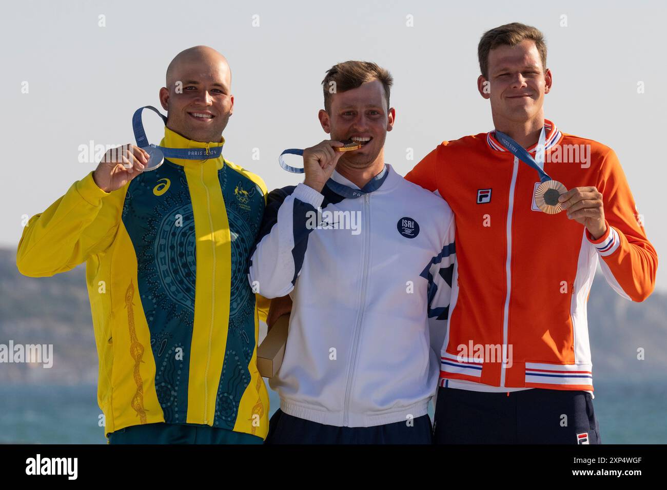 Tom Reuveny of Israel, center, bites his men's iQFOiL windsurfing gold ...