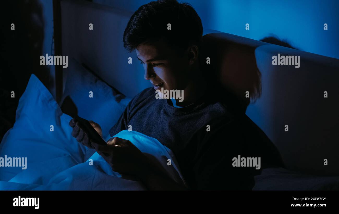 Young man scrolling through his smartphone in bed at night under blue glow of technology ...