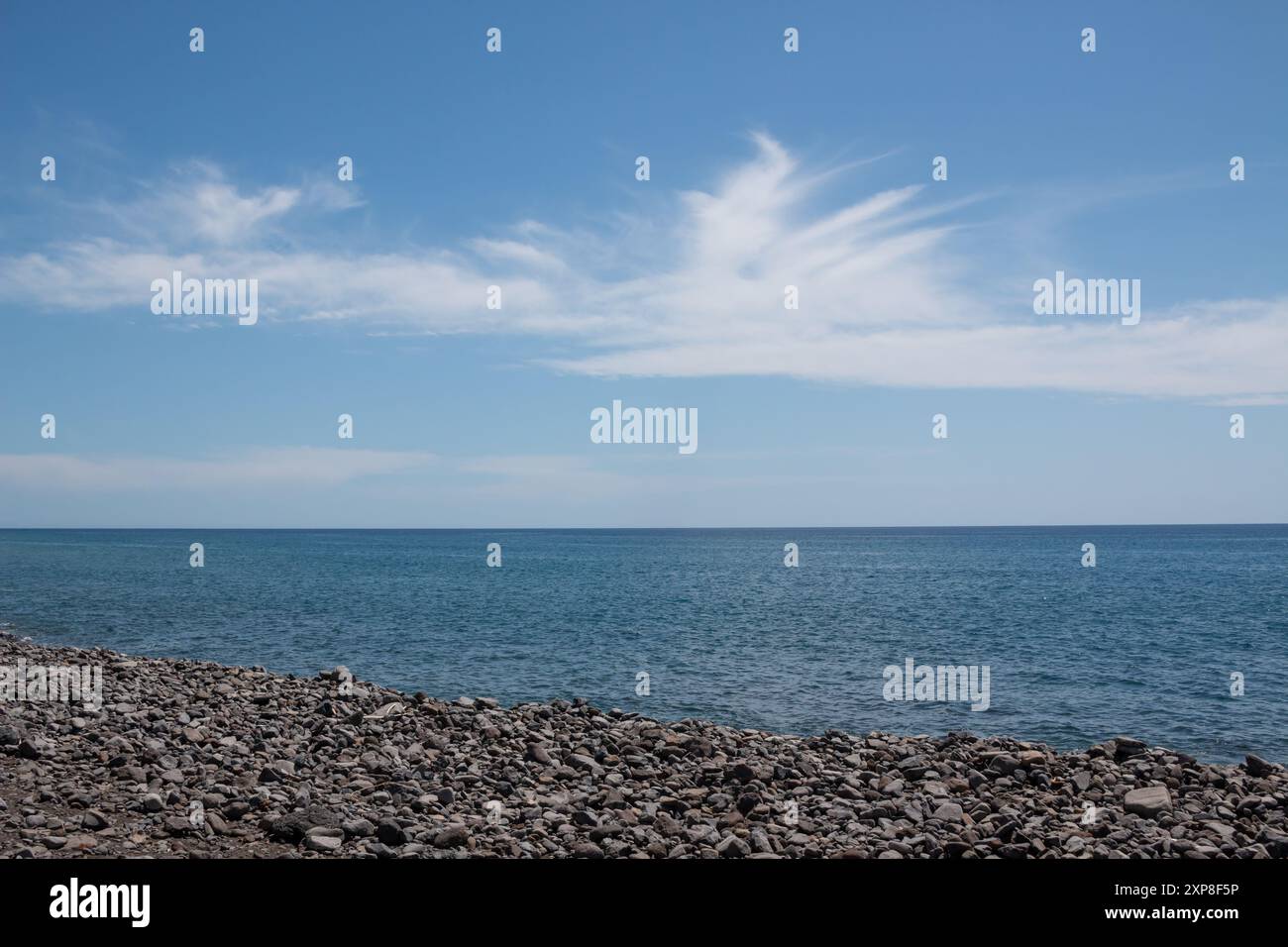 Dark color of the volcanic pebbles on the beach. Blue calm water of ...