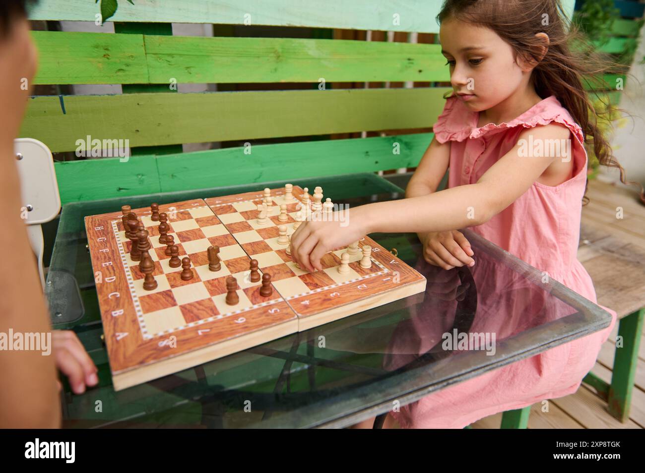 A young girl in a pink dress is playing chess outdoors with a friend ...