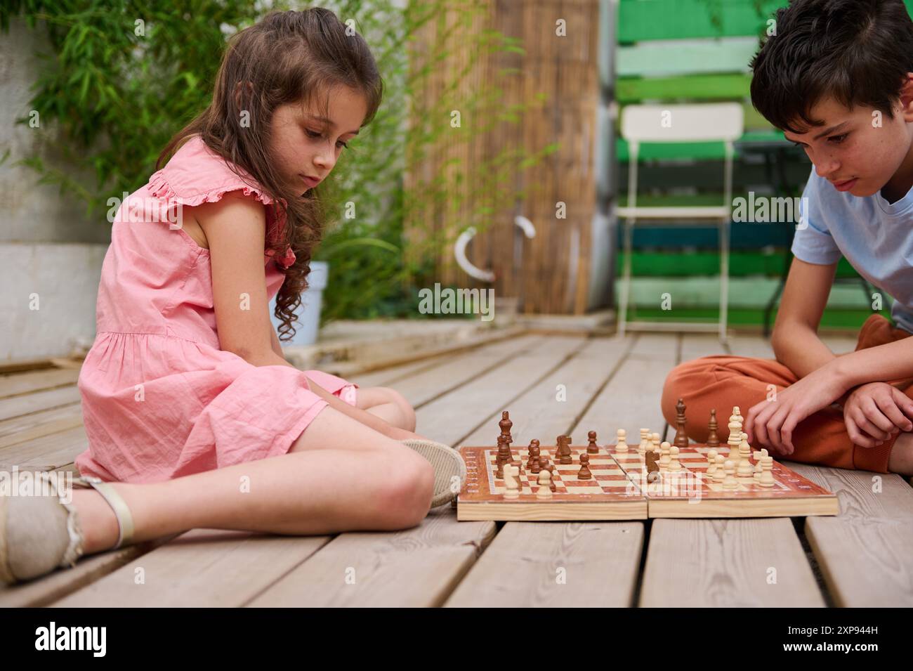 Two children engage in a chess game outdoors on a wooden deck, concentrating on their moves. The ...