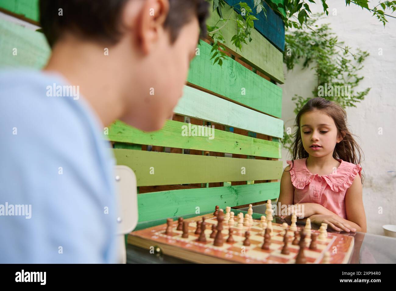 Two kids engaged in a game of chess outside, on a vibrant wooden bench ...