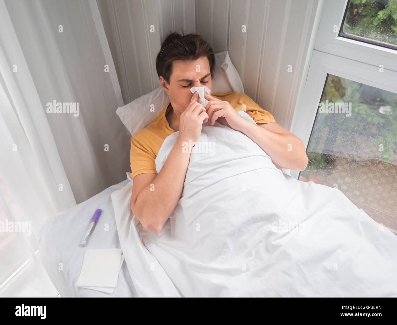 Sick man is lying in bed with thermometer. Man is blowing his nose ...