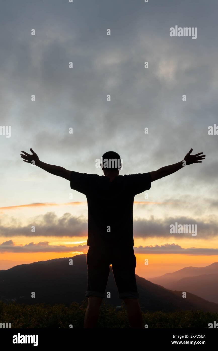 silhouette of young man standing on top of mountain peak with his hands ...
