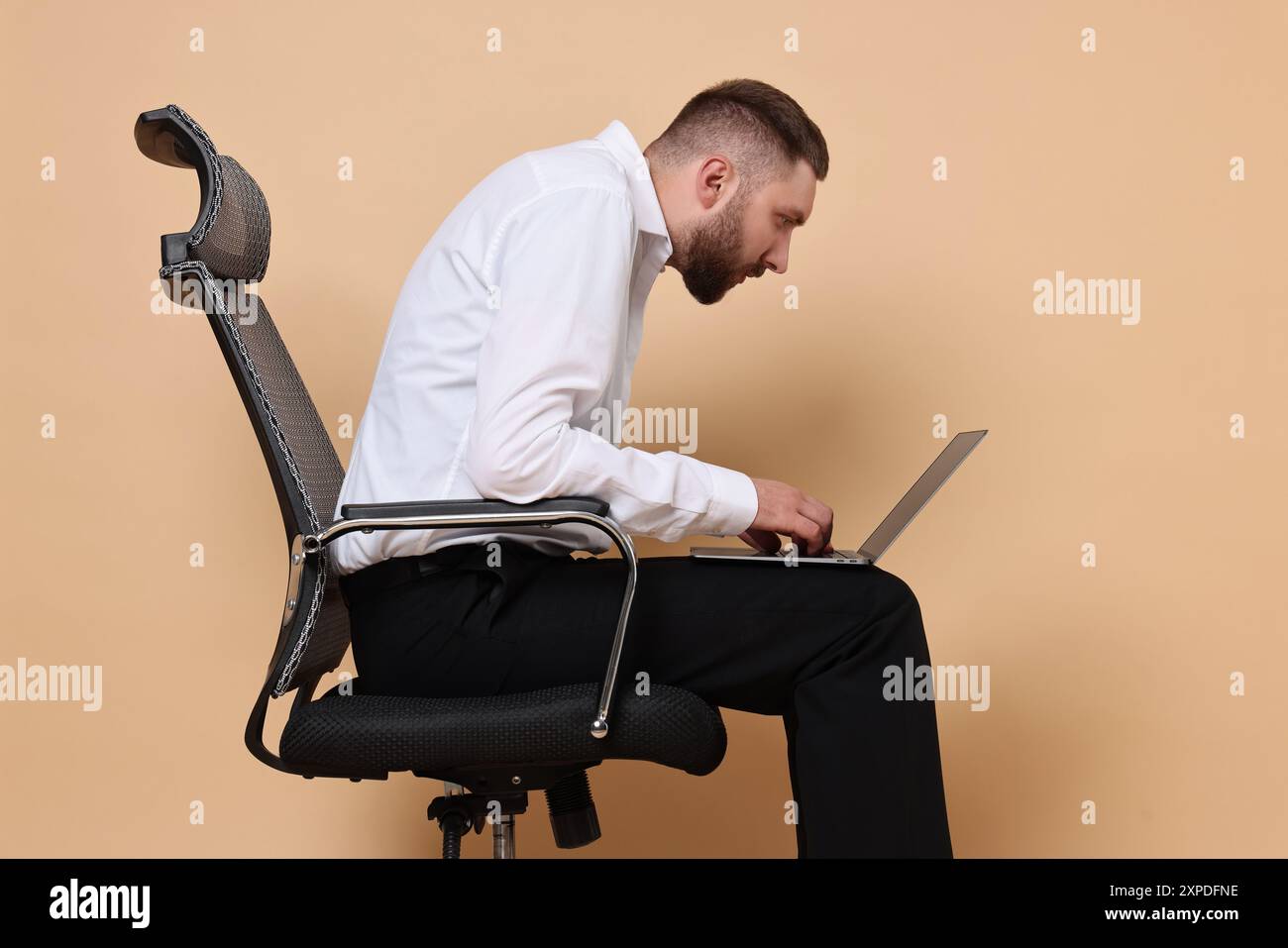 Man with poor posture sitting on chair and using laptop against pale ...
