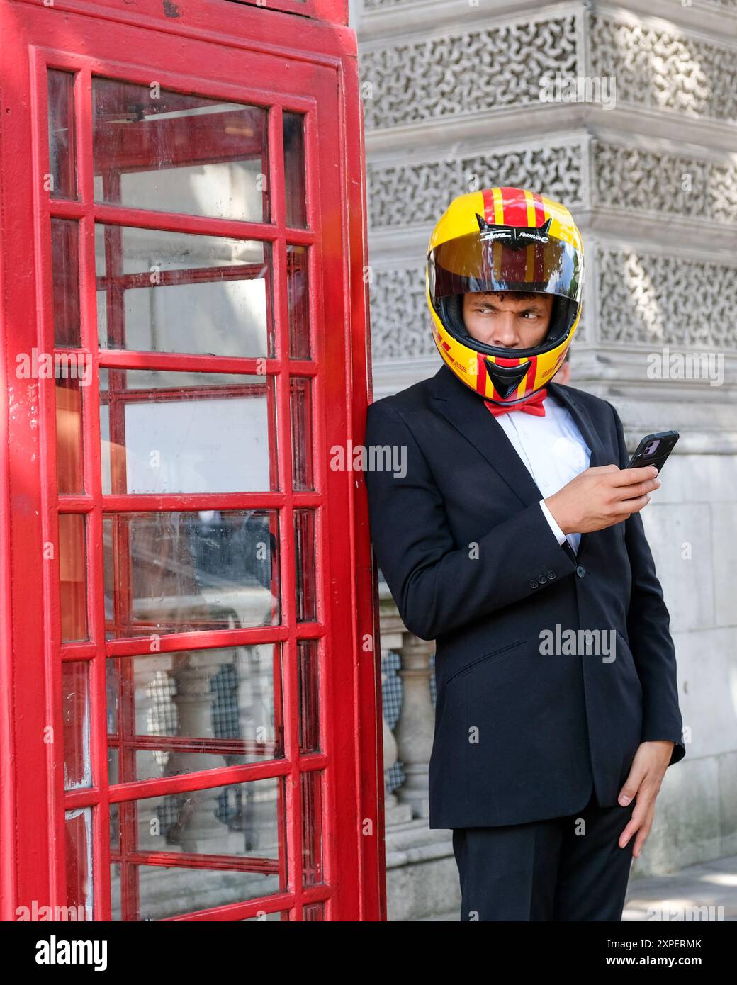A man wearing a tuxedo and crash helmet holding a phone, leans on a red ...