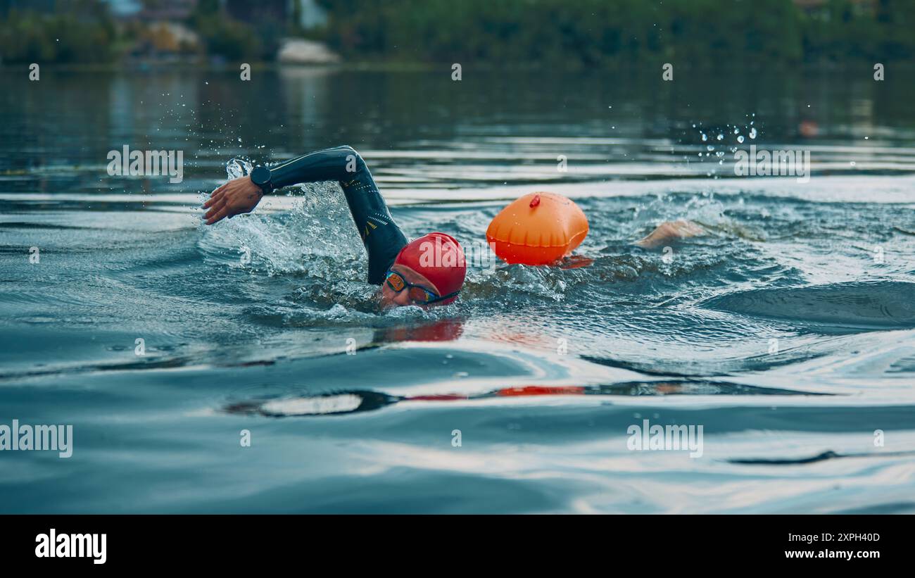Focused swimmer in wetsuit and red cap moves through lake, orange buoy ...