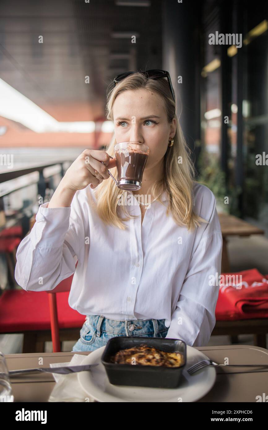 Blonde gen z woman eating lasagna and drink cocoa in the cafe on break. Gen z girl spend time in ...