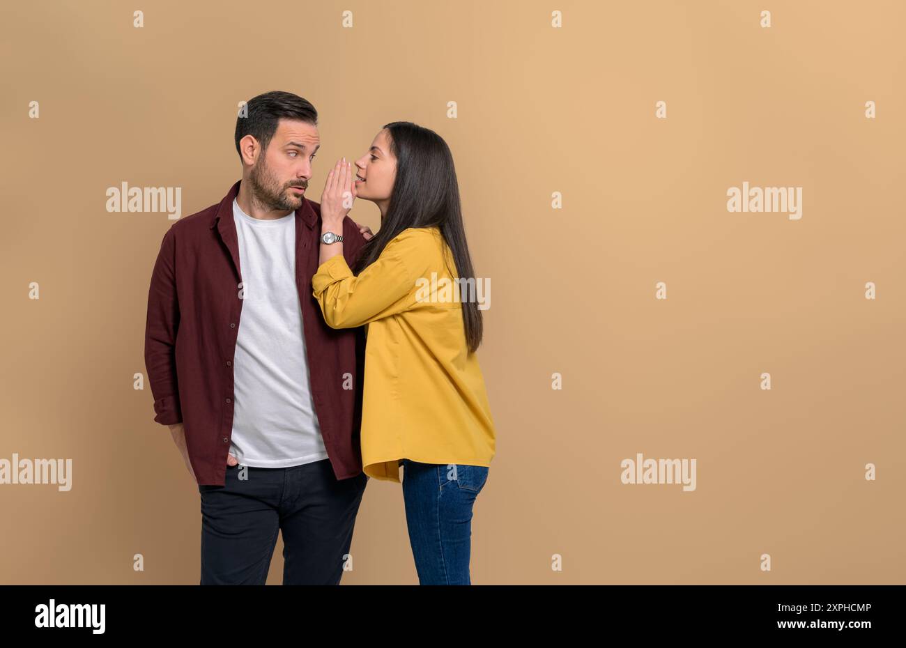 Serious boyfriend looking at girlfriend whispering secret into his ear ...