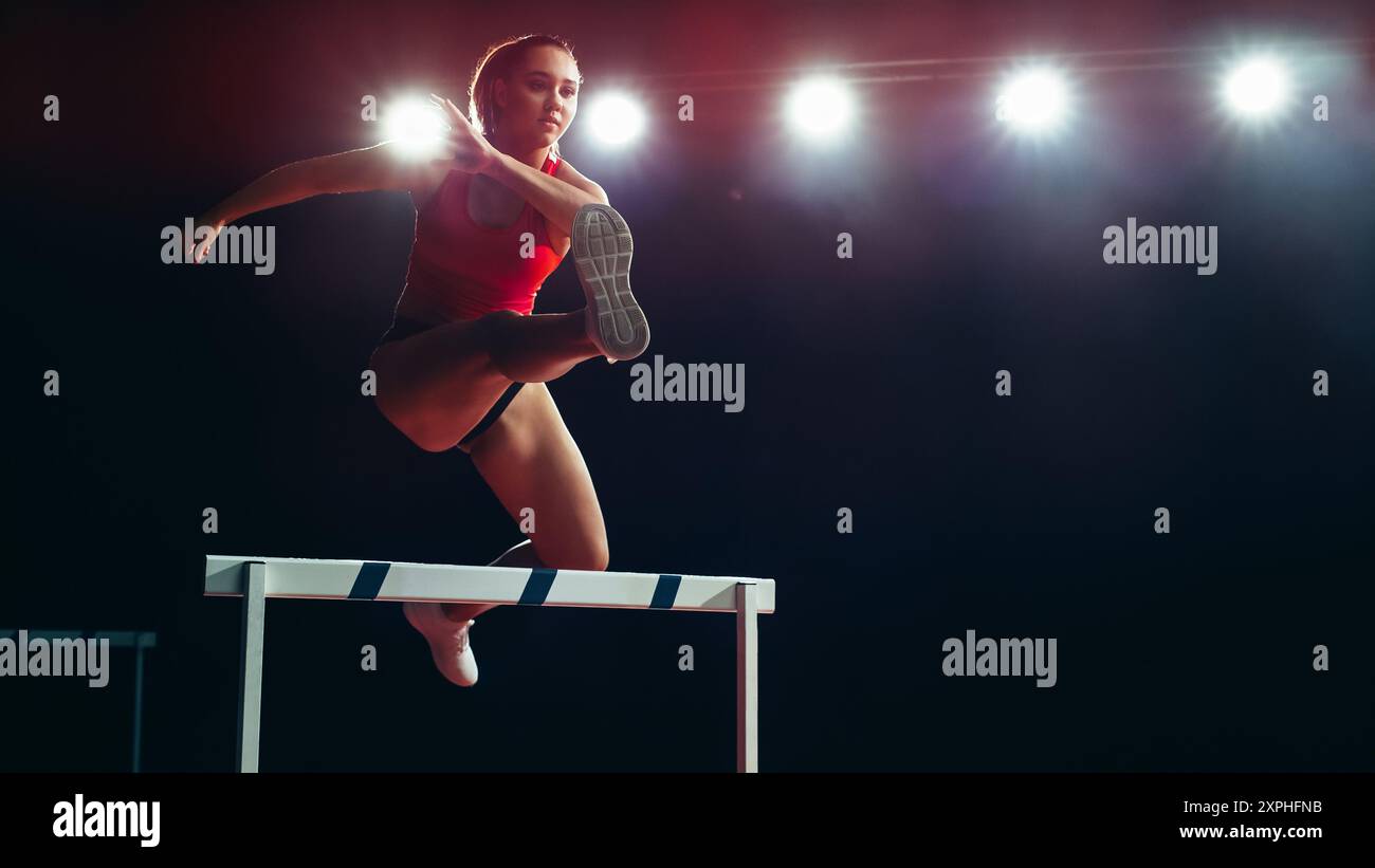 Female Athlete in Mid-air, Leaping Over Hurdle Under Bright Stadium ...