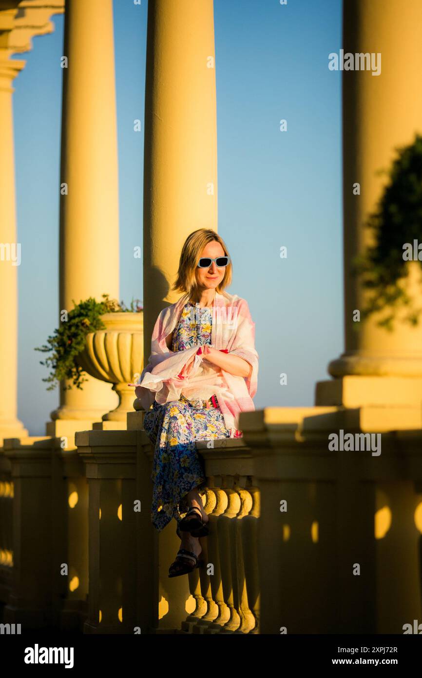 A woman in a summer dress sitting on a parapet in the sunset light ...