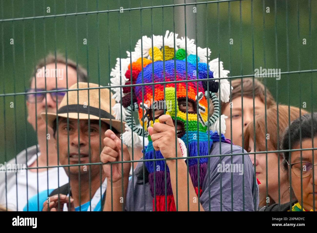 A man wearing a colorful mask looks through a barrier fence as athletes ...