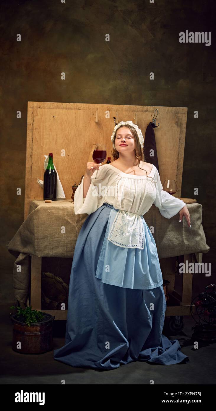 Joyful, young woman in old-fashion dress holding glass of wine standing ...