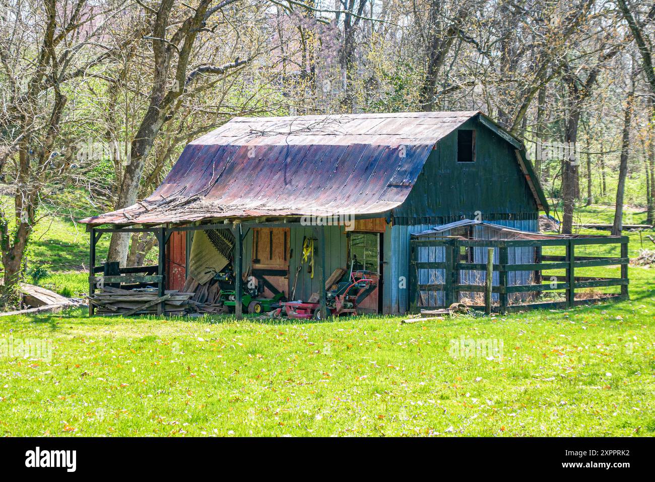 A small, old, and dilapidated barn sits in a grassy field. The barn is surrounded by trees and ...