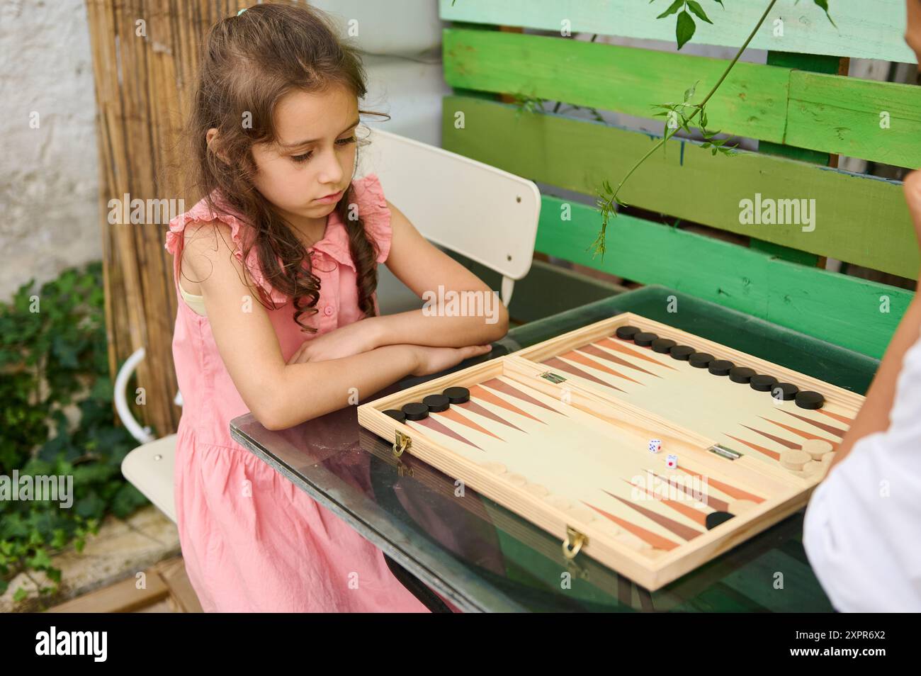 A young girl in a pink dress sits at an outdoor table, playing a game ...