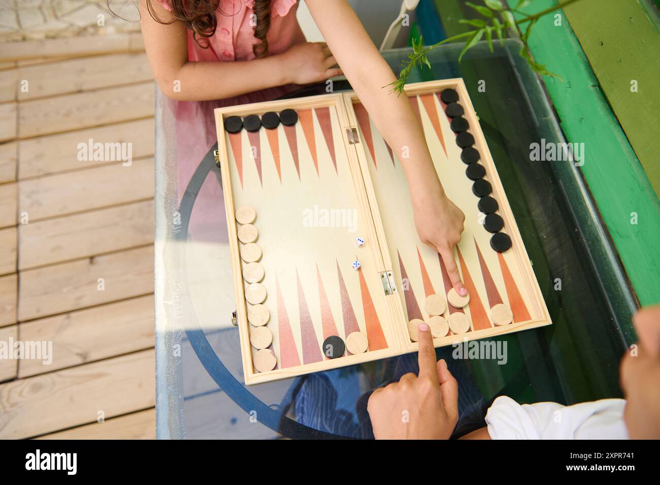 Two children enjoy a game of backgammon on a glass table outside. The image showcases a fun and ...