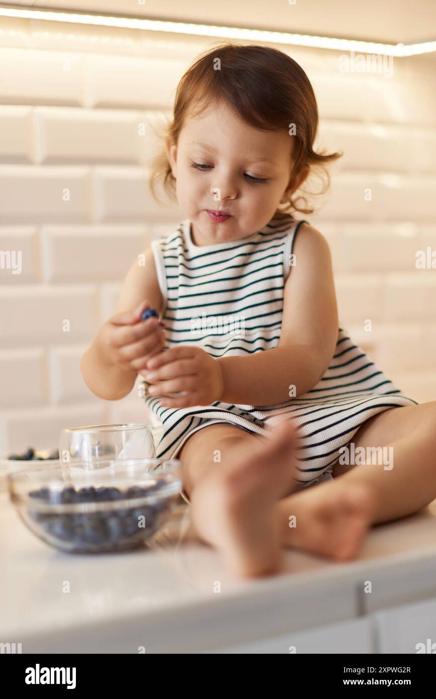 Little baby girl eating fresh blueberries from glass bowl in white ...