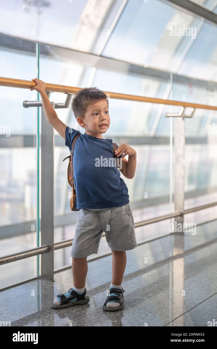 A young boy in a blue shirt and gray shorts, with a backpack, stands ...