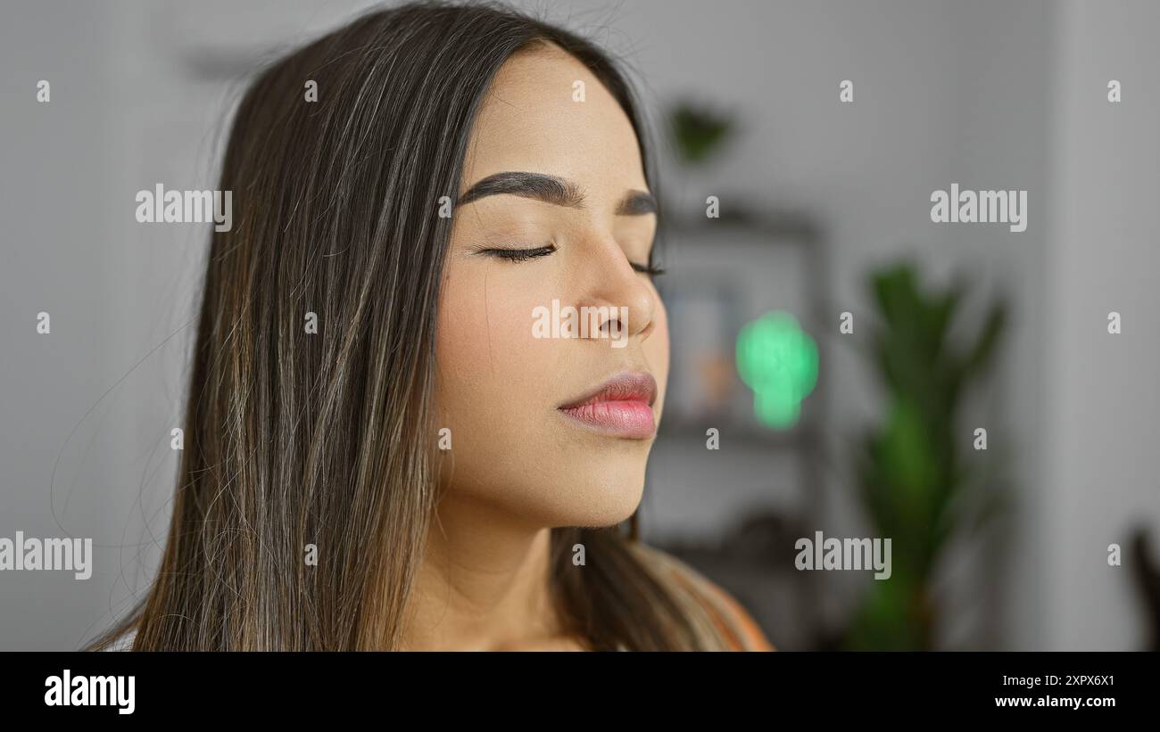 A serene young hispanic woman poses indoors, eyes closed, with a plant ...