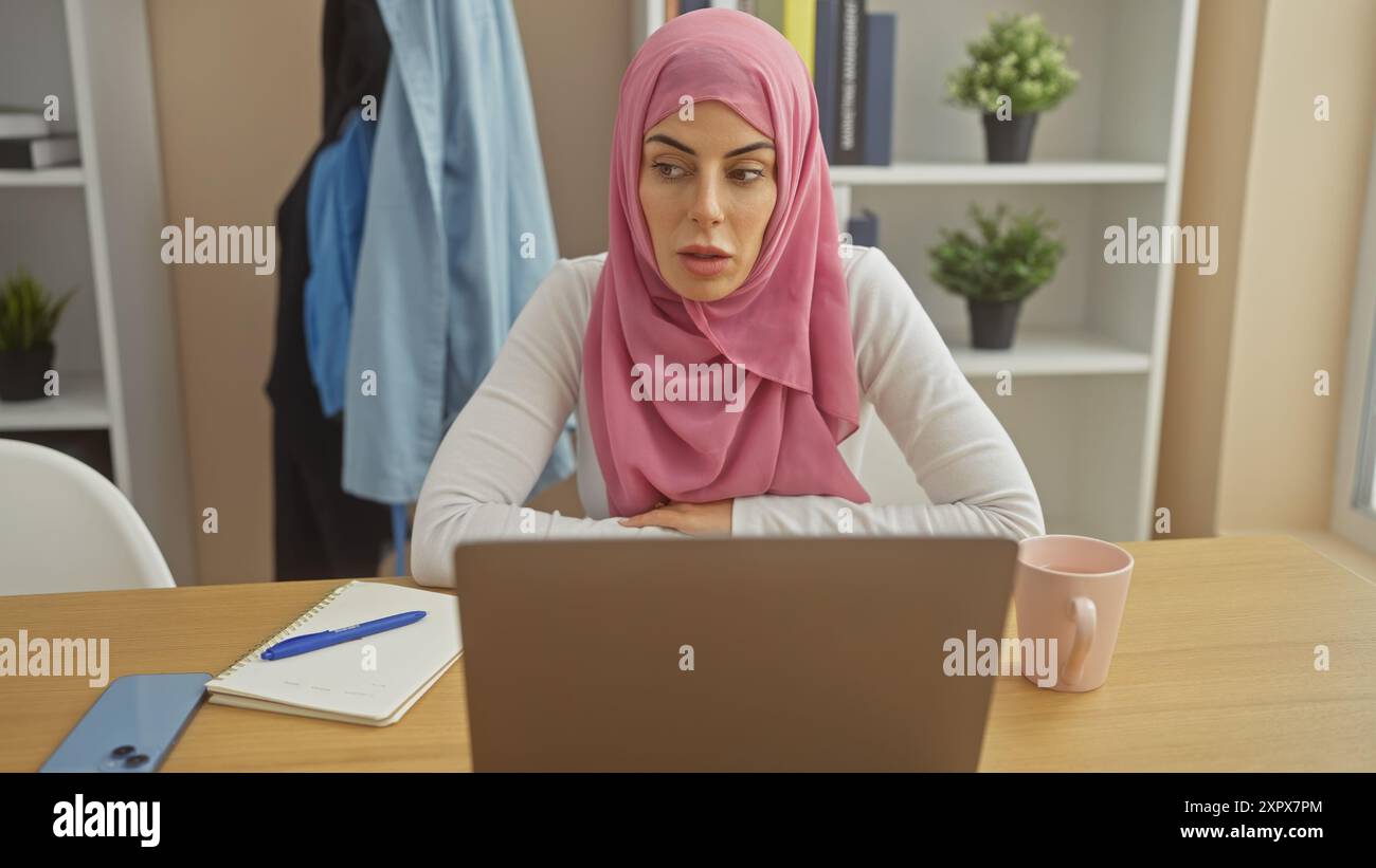 A focused woman wearing a hijab works on a laptop in a modern home ...