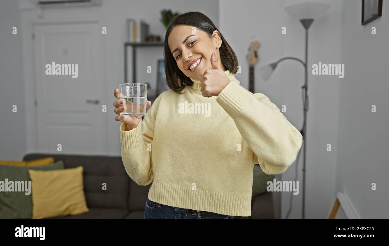 Smiling young hispanic woman holding water glass gives a thumbs up inside a cozy living room ...
