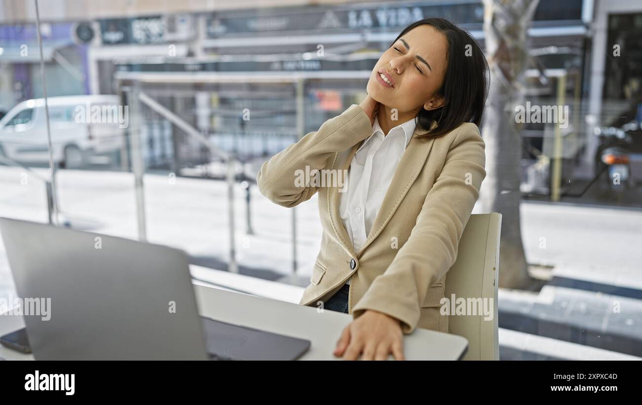 A young hispanic woman in business attire expresses neck pain while ...