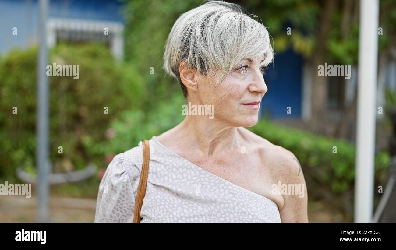 A graceful mature woman with short grey hair stands pensively in a lush ...