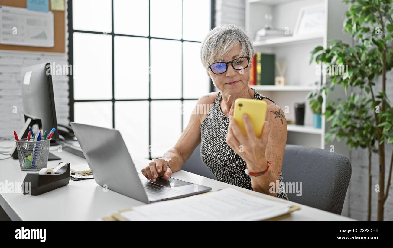 A professional mature woman uses a smartphone and laptop in an office ...